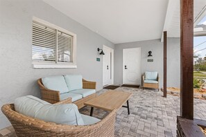 Uma varanda coberta com móveis de vime, uma mesa de madeira e uma porta que dá para o interior.