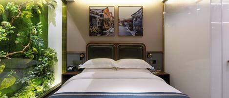 Quarto de hotel com uma cama grande, parede de vidro com parede verde e plantas, duas imagens emolduradas na parede e uma mesa de cabeceira com abajur.