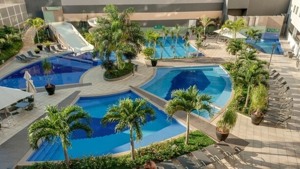 Área de piscina no terraço, com várias piscinas, palmeiras, espreguiçadeiras e design arquitetônico moderno.