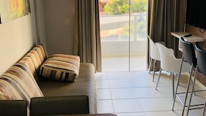 Uma sala de estar moderna com sofá, mesa de jantar e televisão. Há uma porta de vidro deslizante que dá para uma área externa com vista para prédios e vegetação.