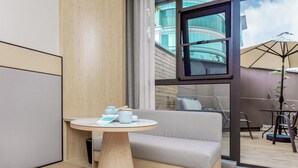 Um interior moderno com uma mesa de madeira redonda, um sofá cinzento e vista para um edifício através de uma porta de vidro.