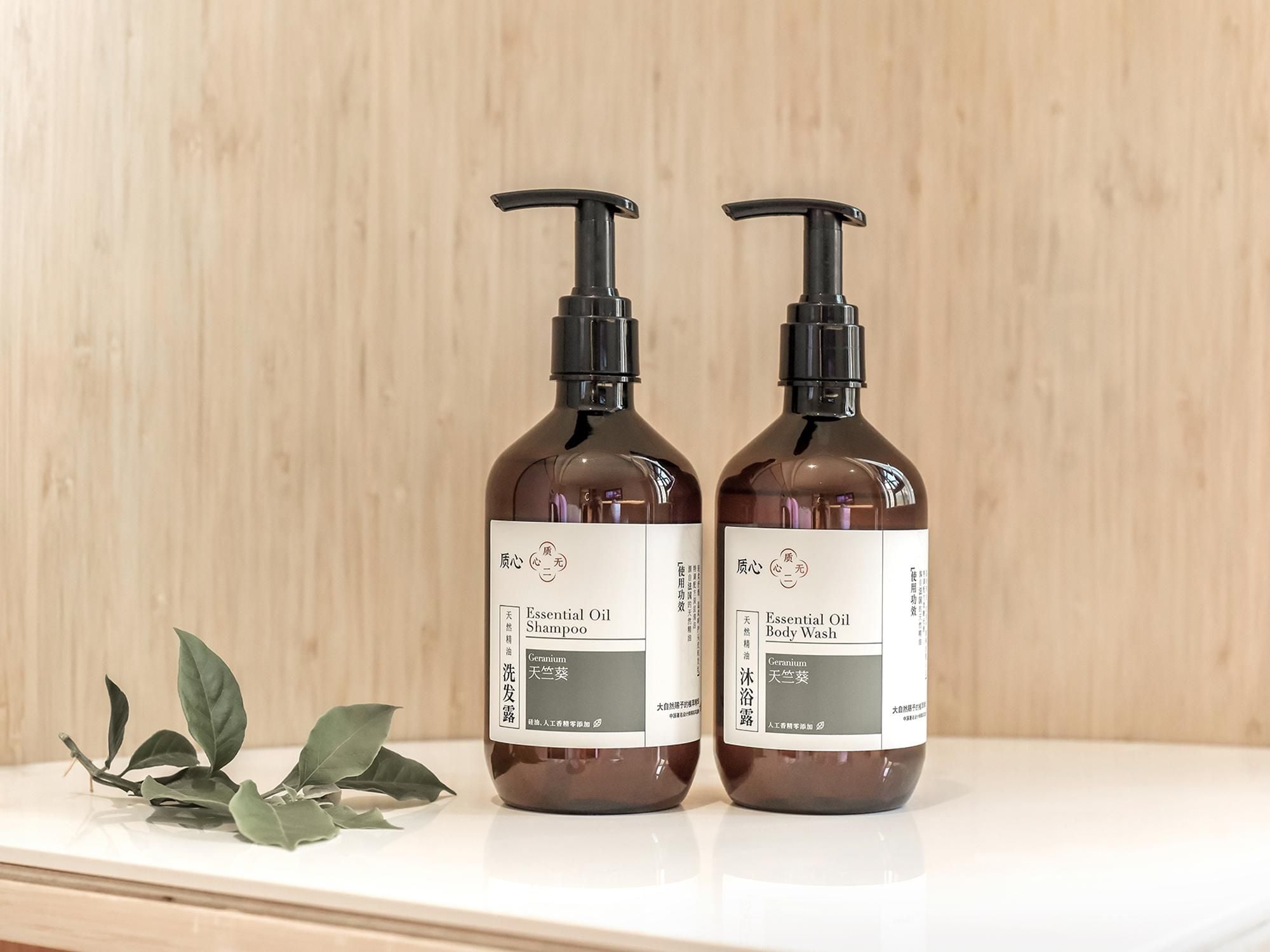 Dois frascos de shampoo e líquido de banho com óleo essencial e bombas, sobre uma superfície branca com fundo de madeira.