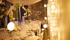 Uma sala de banquetes com mesas redondas dispostas para um evento formal, decorada com flores brancas e velas.