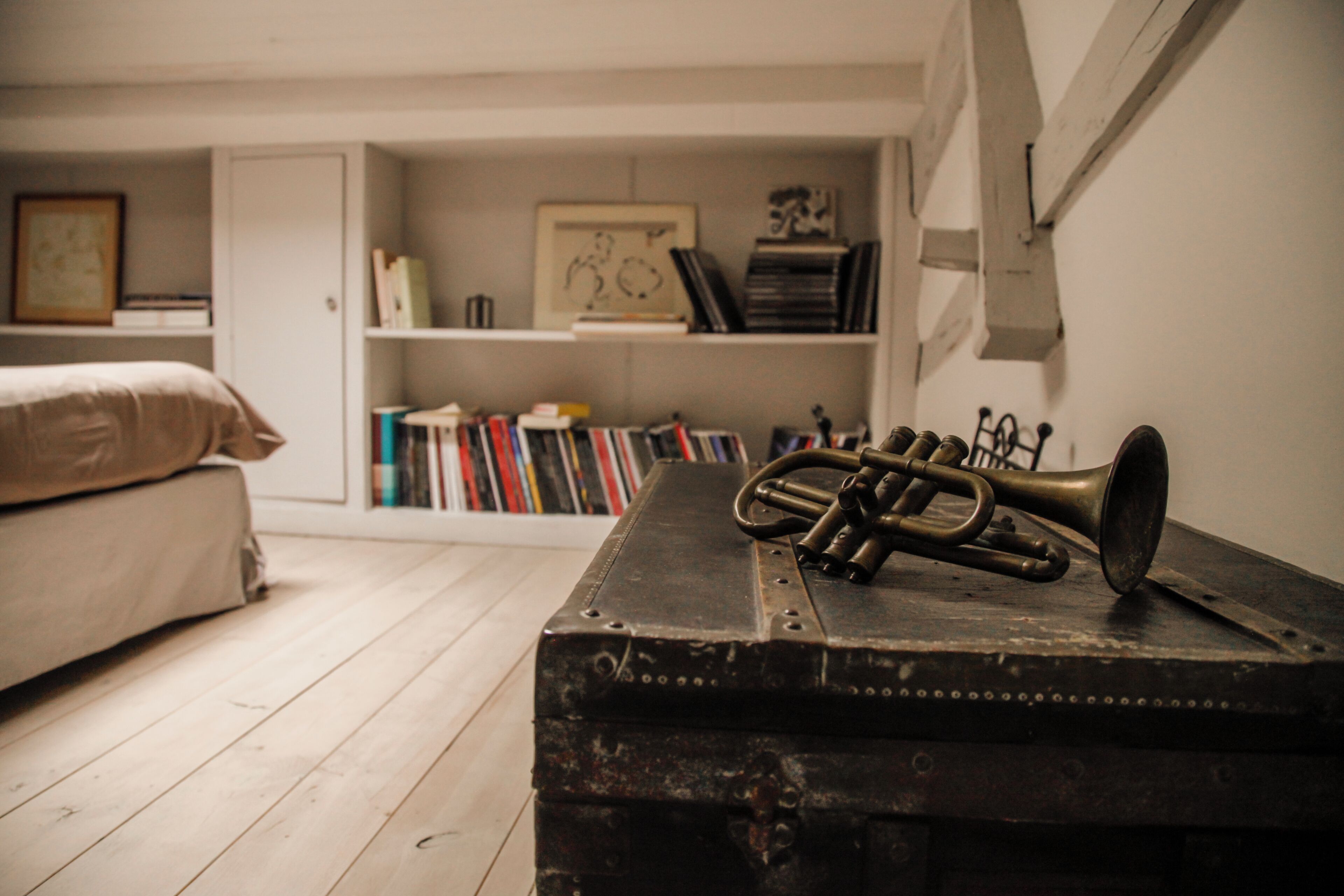Um quarto com cama, piso de madeira, estante com livros e quadros, e uma trompete de latão em um baú de madeira.