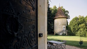Um edifício de pedra com telhado cónico, porta de madeira com detalhes em metal e um espaço de gravilha com uma mesa e cadeiras.