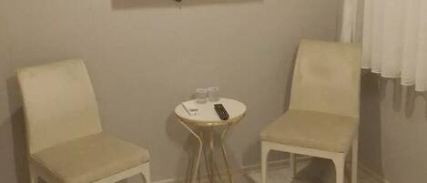 Quarto com duas cadeiras brancas, uma mesa redonda pequena com um controle remoto, e uma TV fixada na parede.