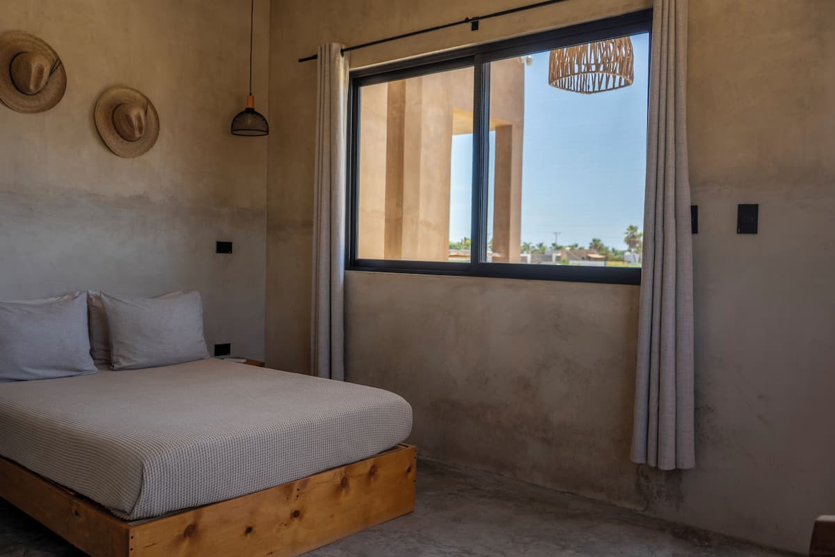 Quarto com cama de madeira, duas almofadas brancas, janela com cortinas e dois chapéus na parede.