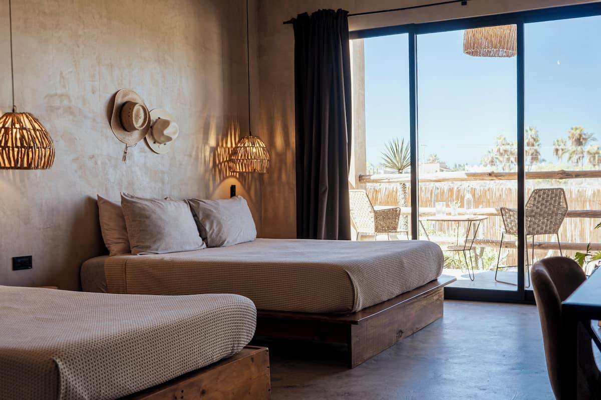 Quarto com uma grande porta de vidro deslizante que dá para um pátio externo. Há duas camas com roupa de cama combinando, uma mesa de madeira e luminárias de vime.