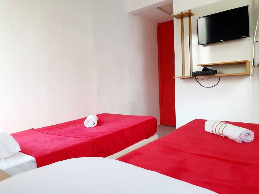 Quarto de hotel com duas camas de solteiro, cada uma com colcha vermelha e toalha branca enrolada. Uma televisão de tela plana está fixada na parede acima de uma prateleira de madeira.