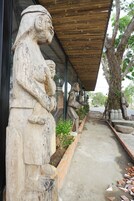 Estátua de pedra de uma figura sentada sob um abrigo de madeira, com uma árvore e um caminho que leva a um edifício.