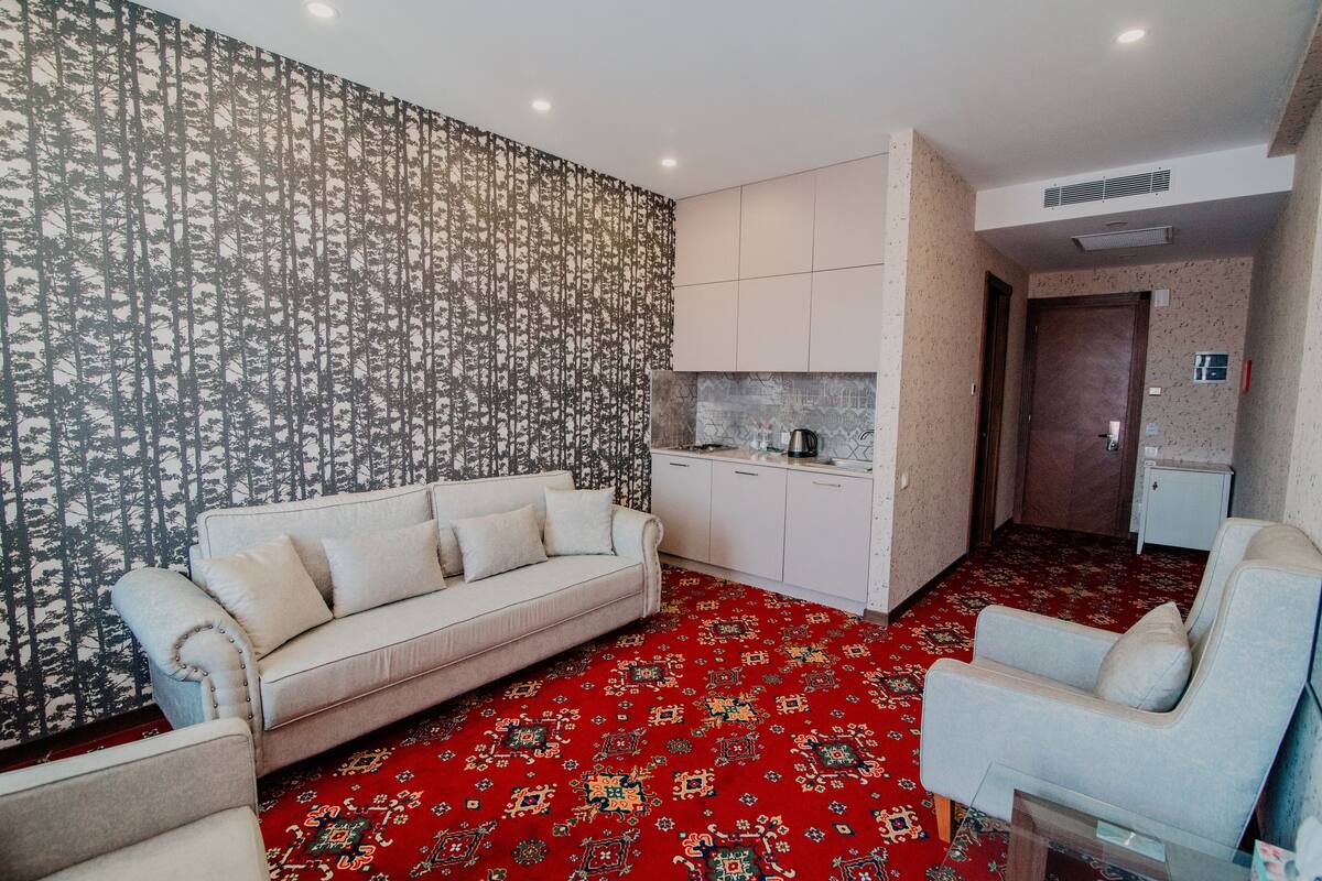 Uma sala de estar com papel de parede estampado, tapete vermelho, sofá, poltrona e um espaço com cozinha compacta.