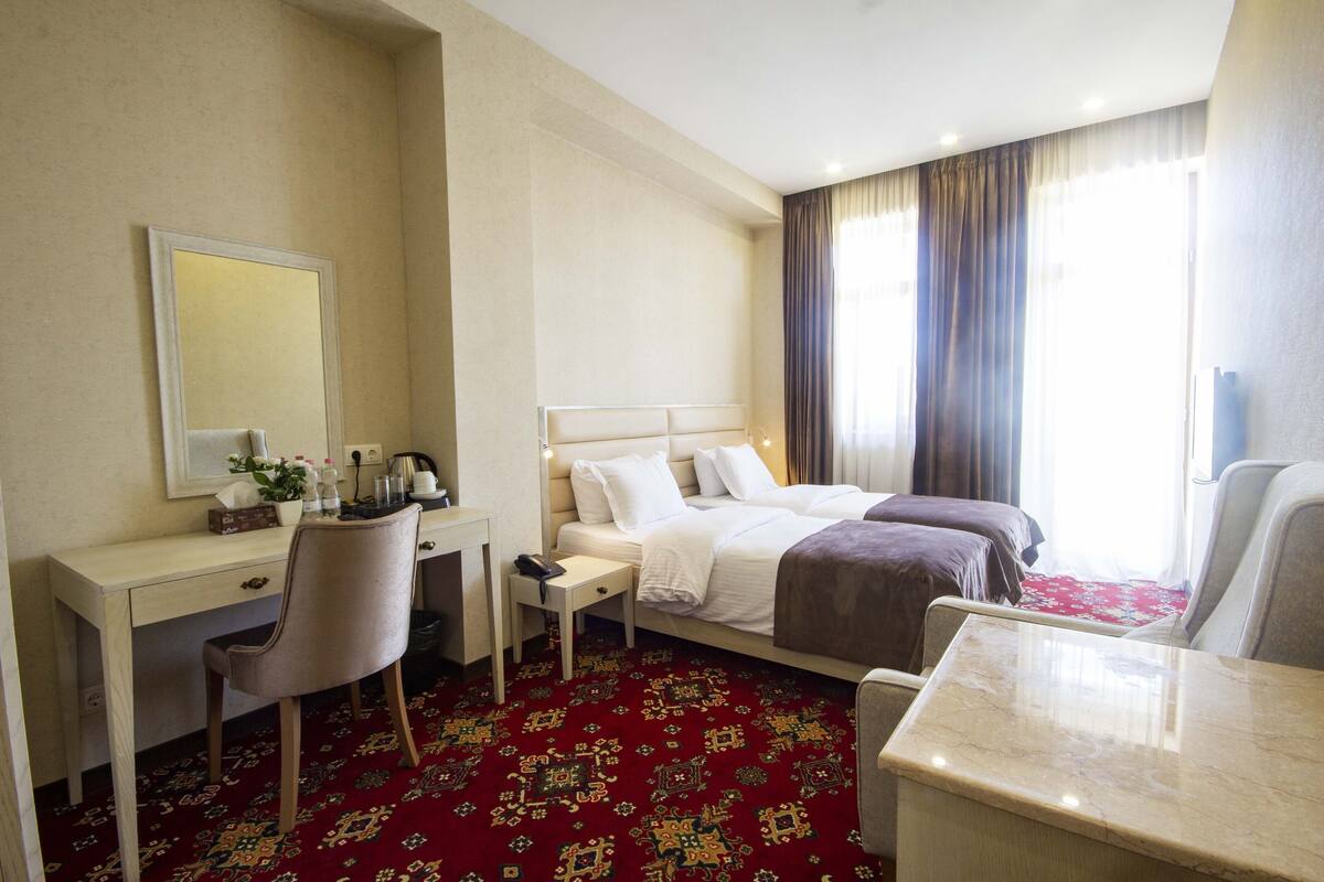 Quarto de hotel com cama, secretária com cadeira, mesa de cabeceira e um espelho grande na parede.