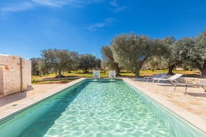Piscina retangular com água cristalina, rodeada por um deck de pedra e espreguiçadeiras.