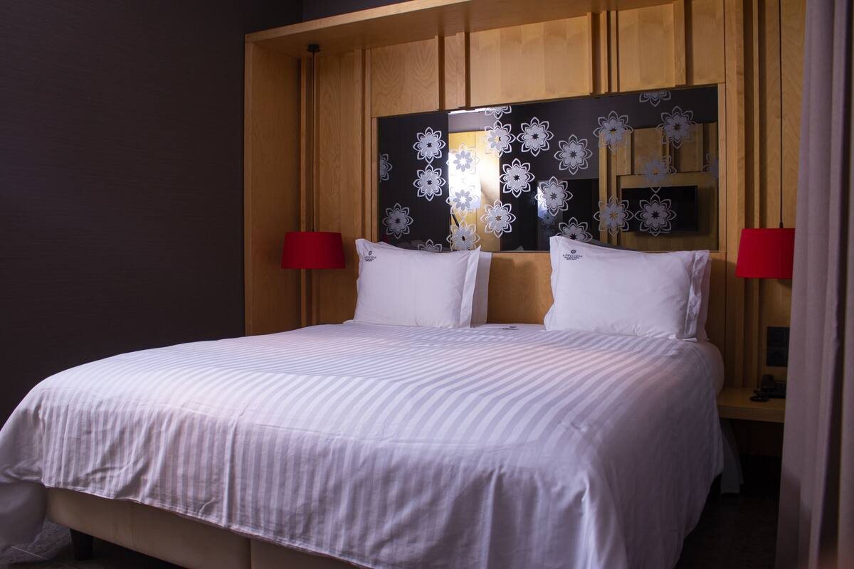 Uma cama de casal com lençóis brancos, duas luminárias laterais vermelhas e um ornamento floral na parede.