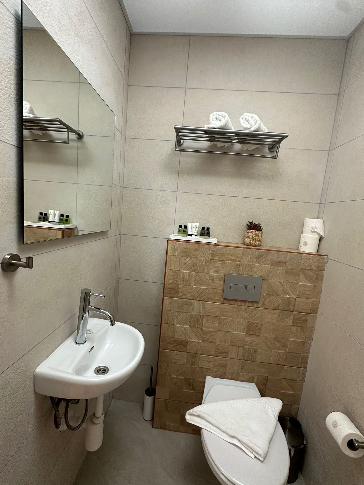 Casa de banho moderna com lavatório branco, sanita e espelho. Há produtos de higiene pessoal na parede e uma toalha sobre a sanita.