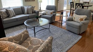Uma sala de estar com sofá cinzento, poltronas, uma mesa de centro e uma área de refeições com mesa e cadeiras.