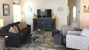 Uma sala de estar com um sofá de cor escura, uma poltrona estampada, uma mesa de centro, uma TV sobre um móvel e um ventilador de teto.