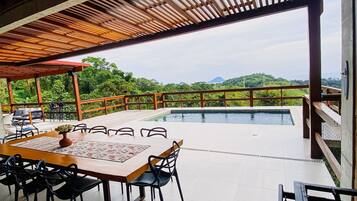 Área de refeições ao ar livre com piscina, pérgola de madeira e vista para a paisagem.