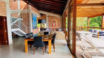 Um espaço amplo e arejado, com teto alto, mesa de jantar com cadeiras, escada e vista através de grandes janelas.