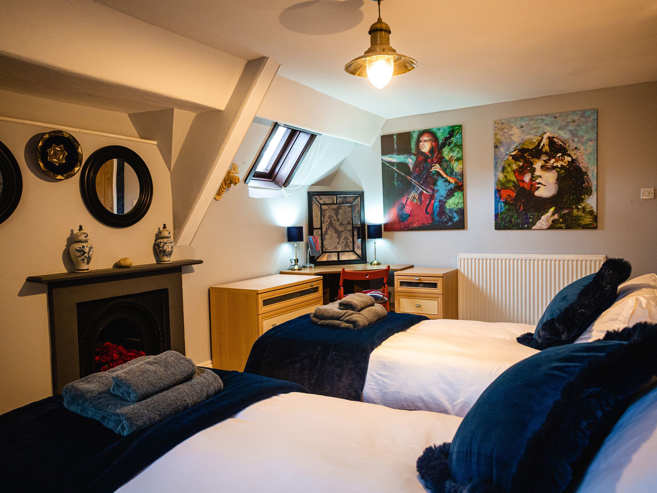 Quarto com cama, mesas de cabeceira, escrivaninha, cadeira, lareira e obras de arte nas paredes.