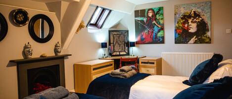 Quarto com cama, mesas de cabeceira, escrivaninha, cadeira, lareira e obras de arte nas paredes.