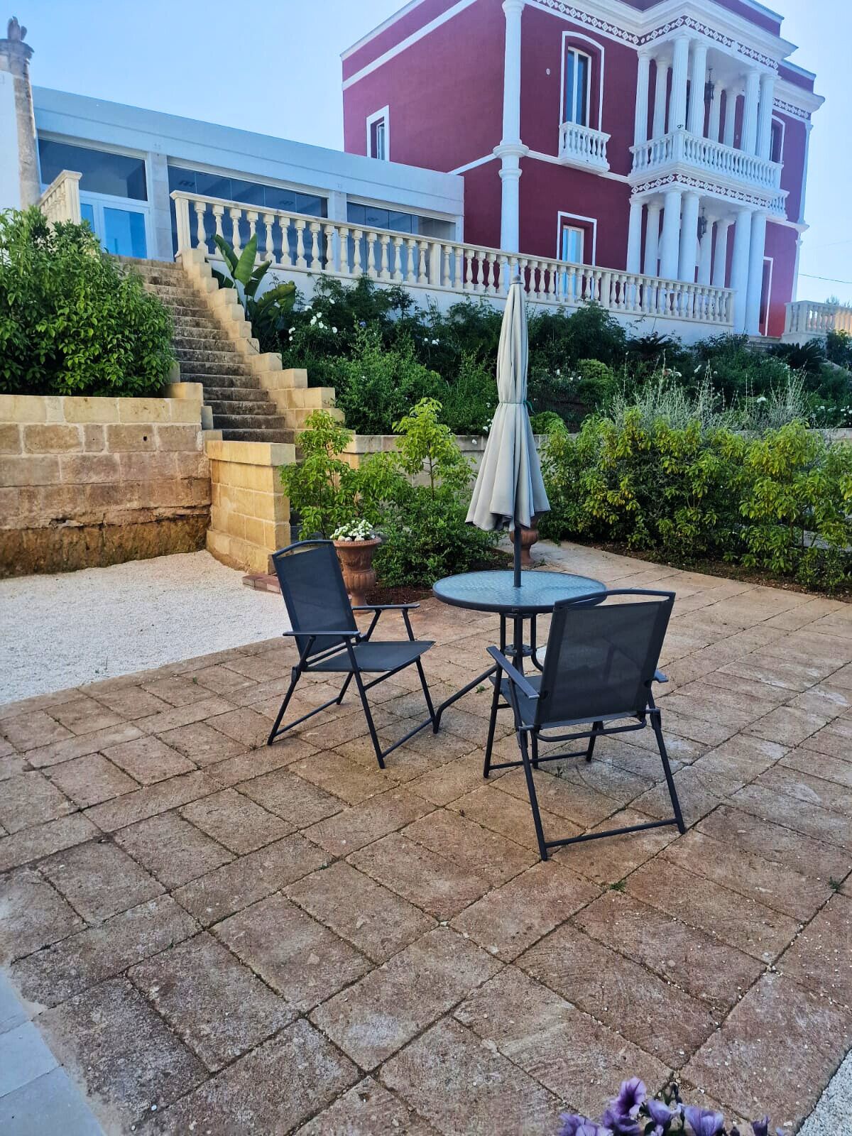 Pátio com duas cadeiras e uma mesa, cercado por um muro de pedra e vegetação.