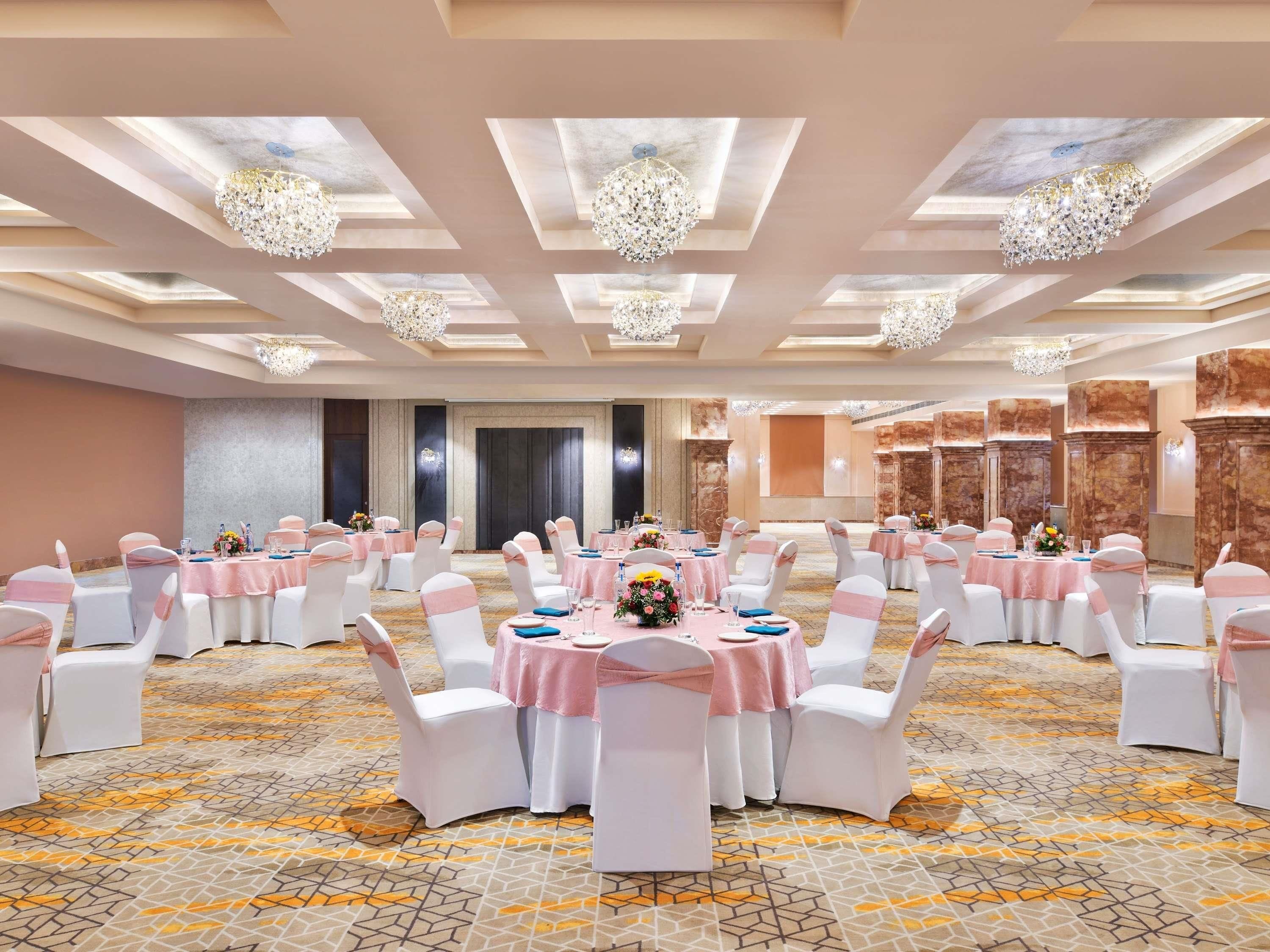 Uma sala de banquetes com mesas redondas, cadeiras brancas e lustres.
