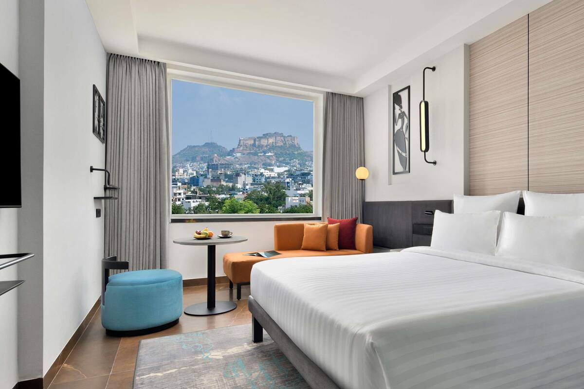 Quarto de hotel com uma cama grande, área de estar, uma mesinha com frutas, televisão e vista para a paisagem urbana.