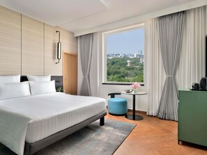 Quarto de hotel com uma cama grande, uma mesinha com um vaso de flores, um armário verde e uma janela com vista para a cidade.