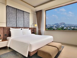 Quarto de hotel moderno com uma cama grande, mesa de cabeceira, luminária e vista para a cidade.