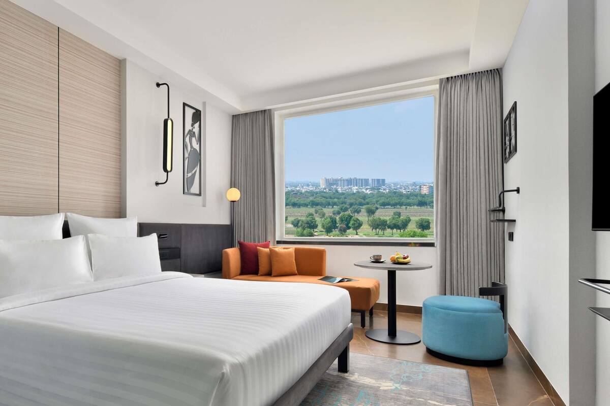 Quarto de hotel com uma cama grande, um sofá, uma mesinha, uma televisão e uma janela com vista para a cidade.