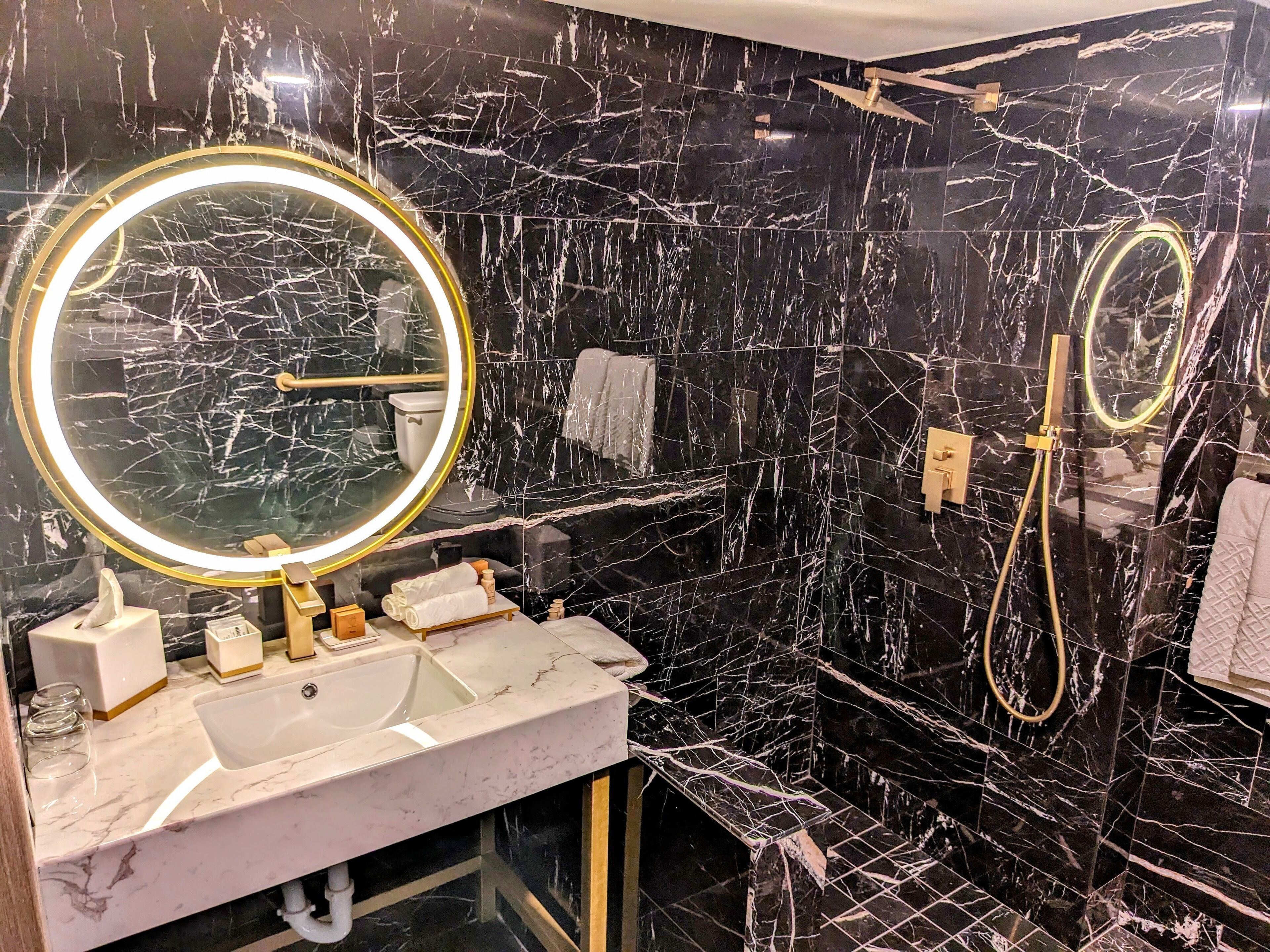 Casa de banho com lavatório de mármore, espelho redondo com moldura dourada, chuveiro manual e porta-toalhas.
