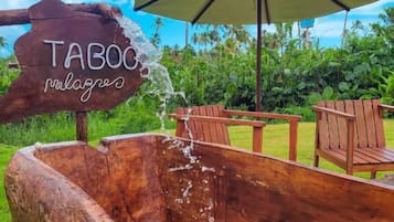 Uma banheira de madeira com água jorrando, cadeiras de madeira e um letreiro que diz "TABOC palaces" sob um grande guarda-chuva.