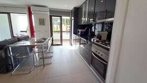 Uma cozinha moderna com armários escuros, uma área de refeições com mesa e cadeiras de vidro e uma vista para uma varanda através de uma porta de correr.