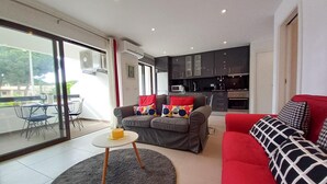 Uma sala moderna com um sofá cinzento, um sofá vermelho e uma mesa de centro redonda. Há uma varanda com uma mesa e cadeiras.