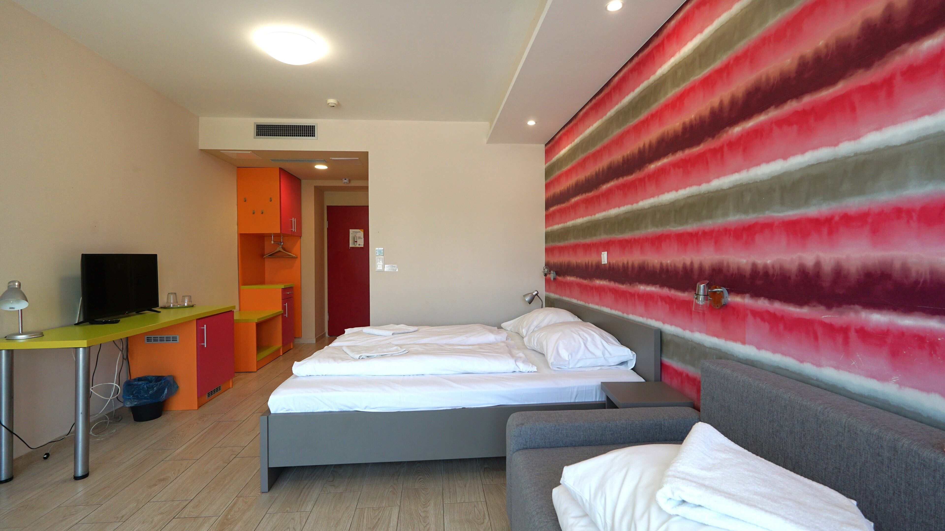 Quarto de hotel com cama, sofá, escrivaninha e parede com padrão listrado vibrante.