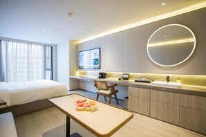 Quarto de hotel moderno com uma cama, uma mesinha com frutas, uma escrivaninha com computador, um espelho e TV na parede.