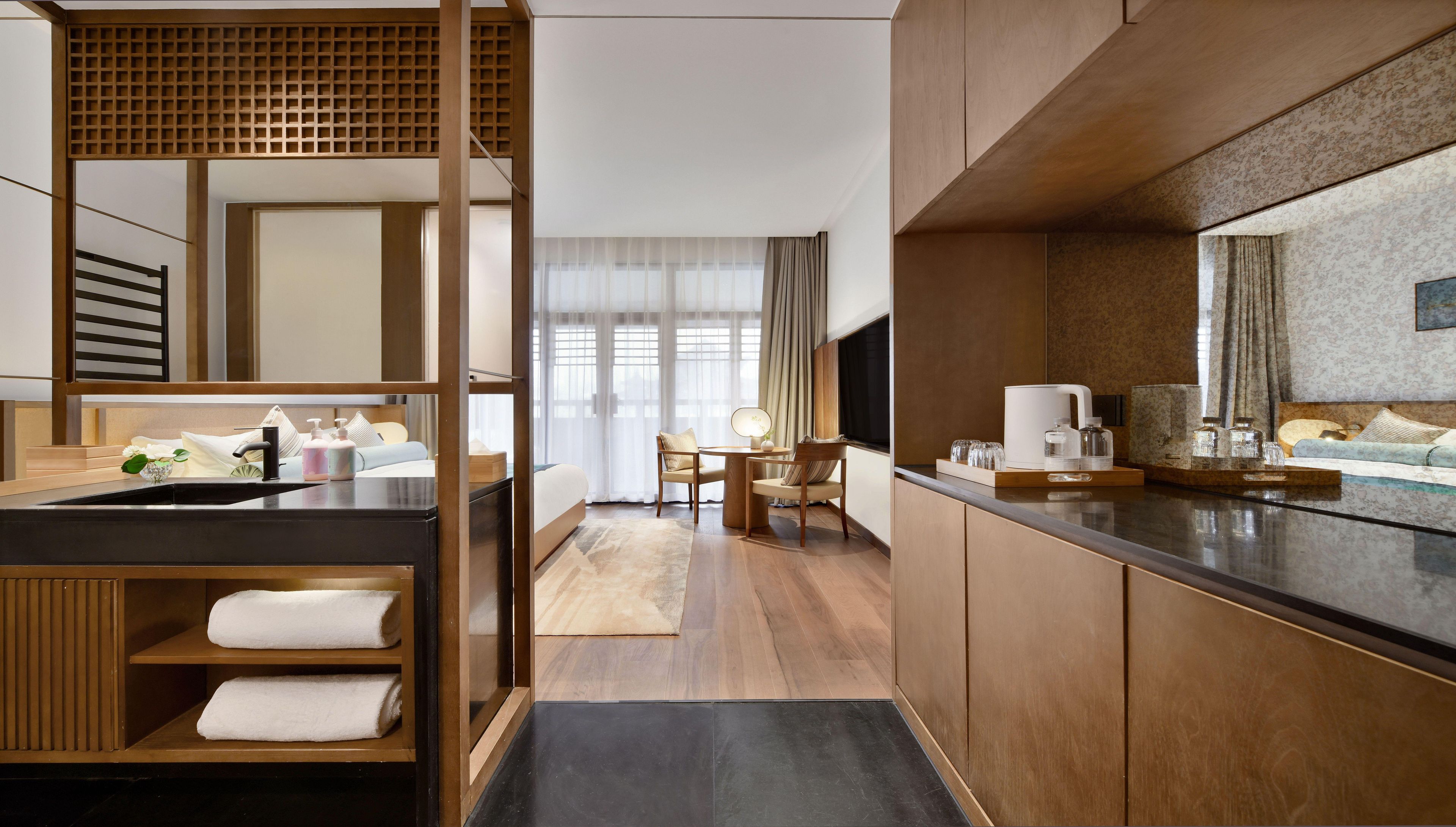 Quarto de hotel com piso de madeira, cama com roupa de cama branca, área de estar com mesa e cadeiras, cozinha compacta com cafeteira e uma janela grande com cortinas.