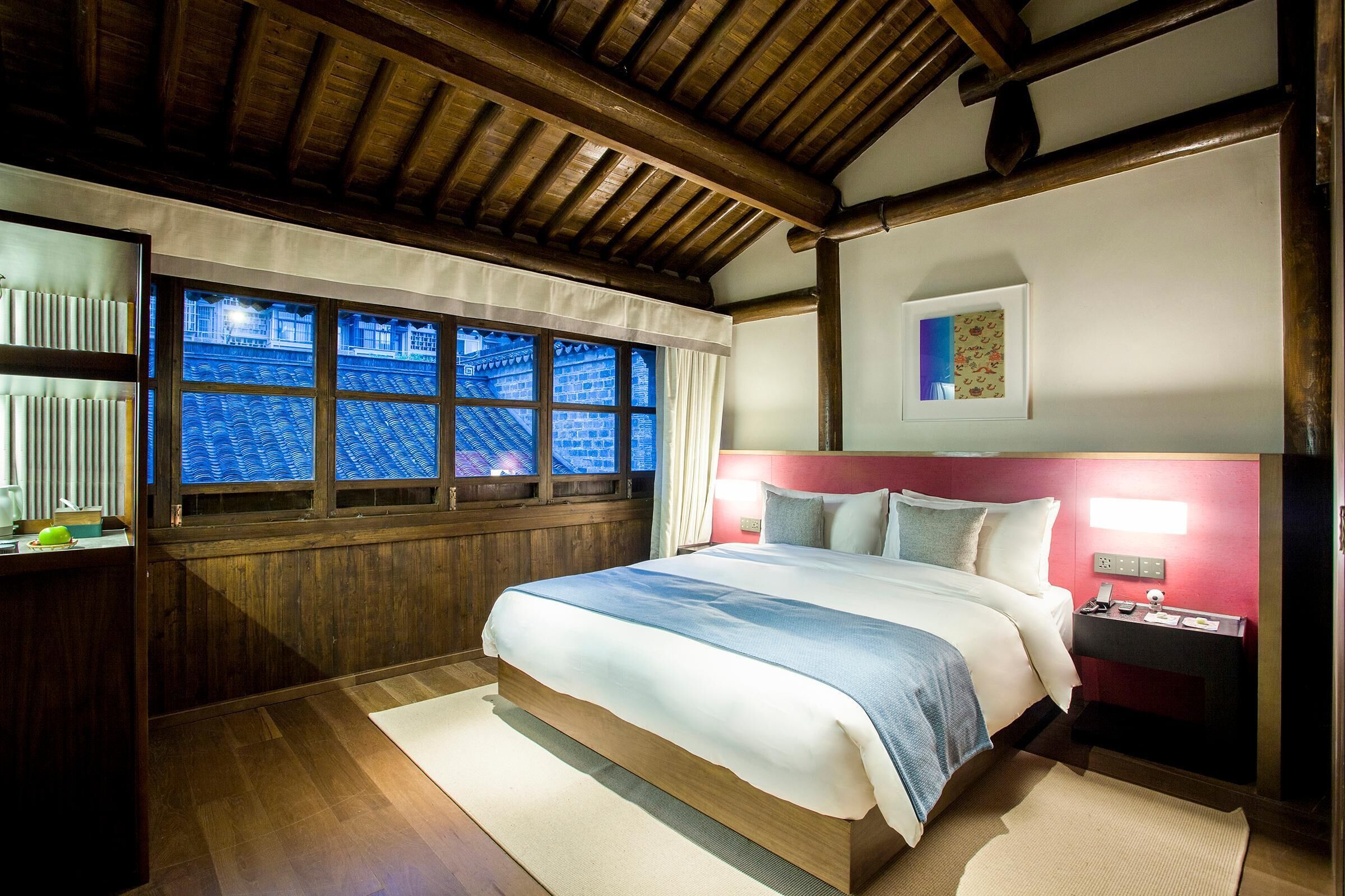 Quarto de hotel com uma cama grande, vigas de madeira no teto, uma janela com vista para um edifício azul e uma mesa de cabeceira com um abajur.