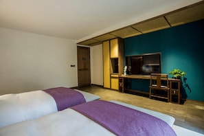 Quarto de hotel com duas camas, uma escrivaninha com cadeira, televisão e uma pequena mesa com planta.