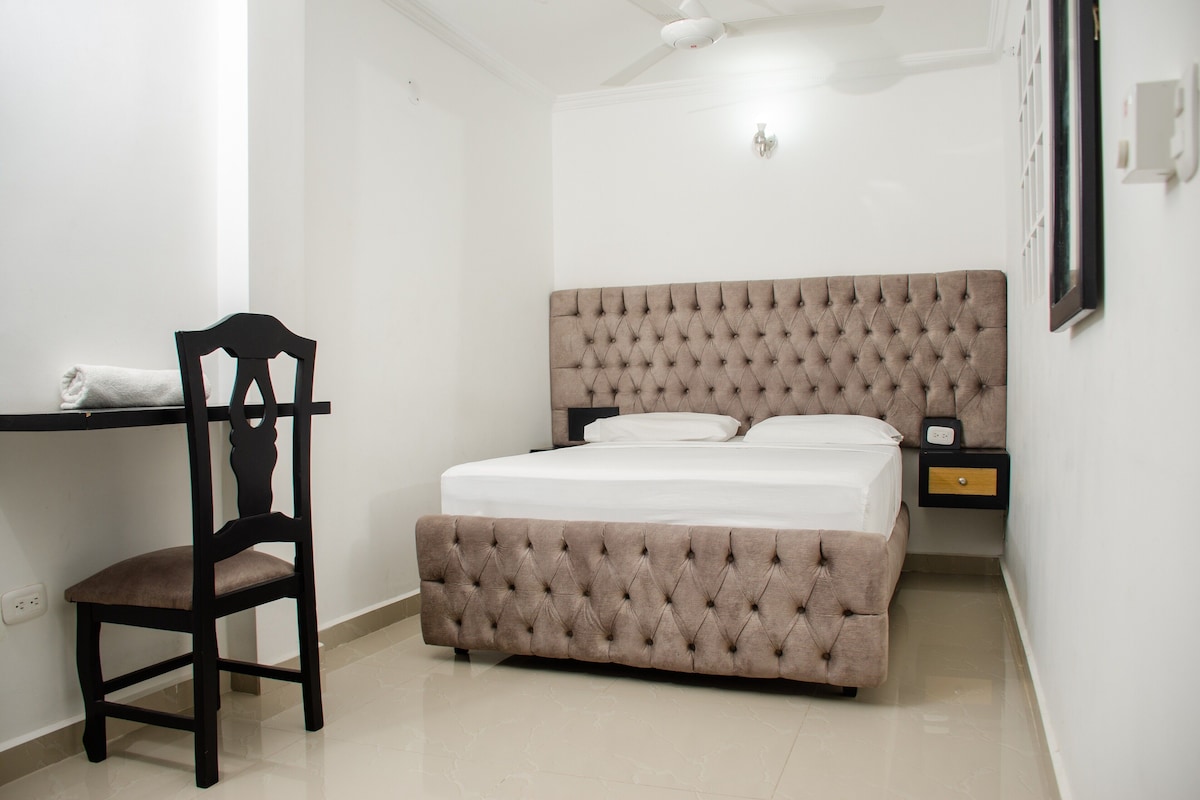 Quarto com cama de cabeceira estofada, uma cadeira de madeira, mesa de cabeceira e criado-mudo.