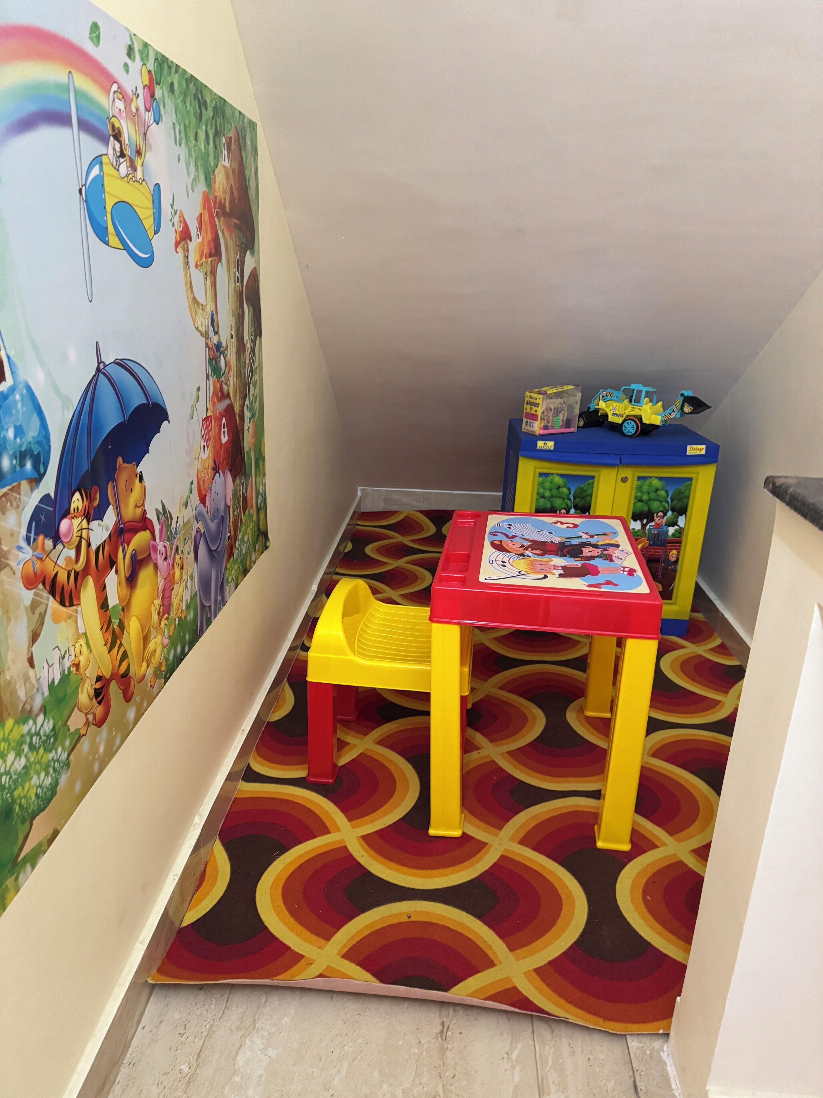 Um espaço infantil com um mural colorido, uma mesinha, uma cadeira amarela e um brinquedo de construção sobre um tapete estampado.