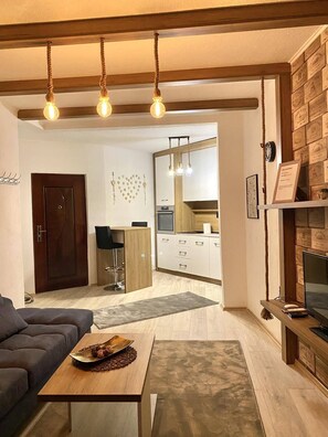 Uma sala de estar moderna com um sofá escuro, uma mesa de centro de madeira e um rack de TV com uma televisão de tela plana. Há uma área de cozinha com armários de madeira e uma mesa de jantar.