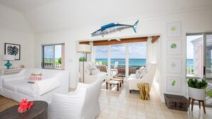 Uma sala de estar espaçosa com uma grande janela com vista para o oceano, mobiliada com sofás brancos e uma mesa de centro de madeira.