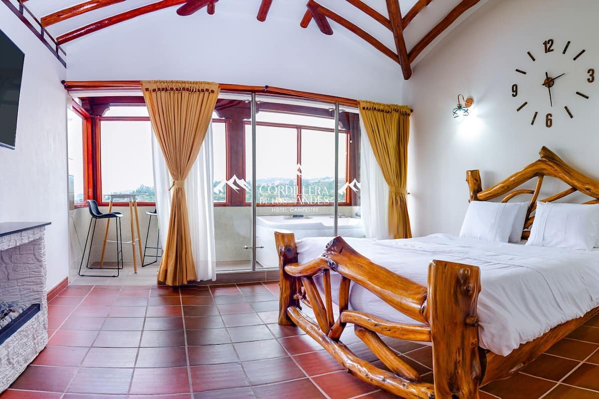 Uma cama de madeira, um relógio, uma mesinha com abajur e vista para o exterior pela janela.
