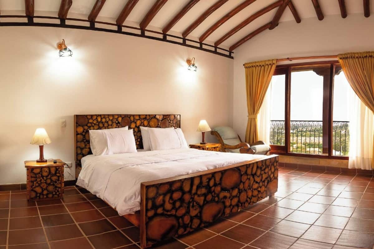 Quarto com cabeceira de pedra, cama com roupa de cama branca, duas luminárias de cabeceira, mesa de cabeceira, cadeira de madeira, varanda com vista e chão ladrilhado.