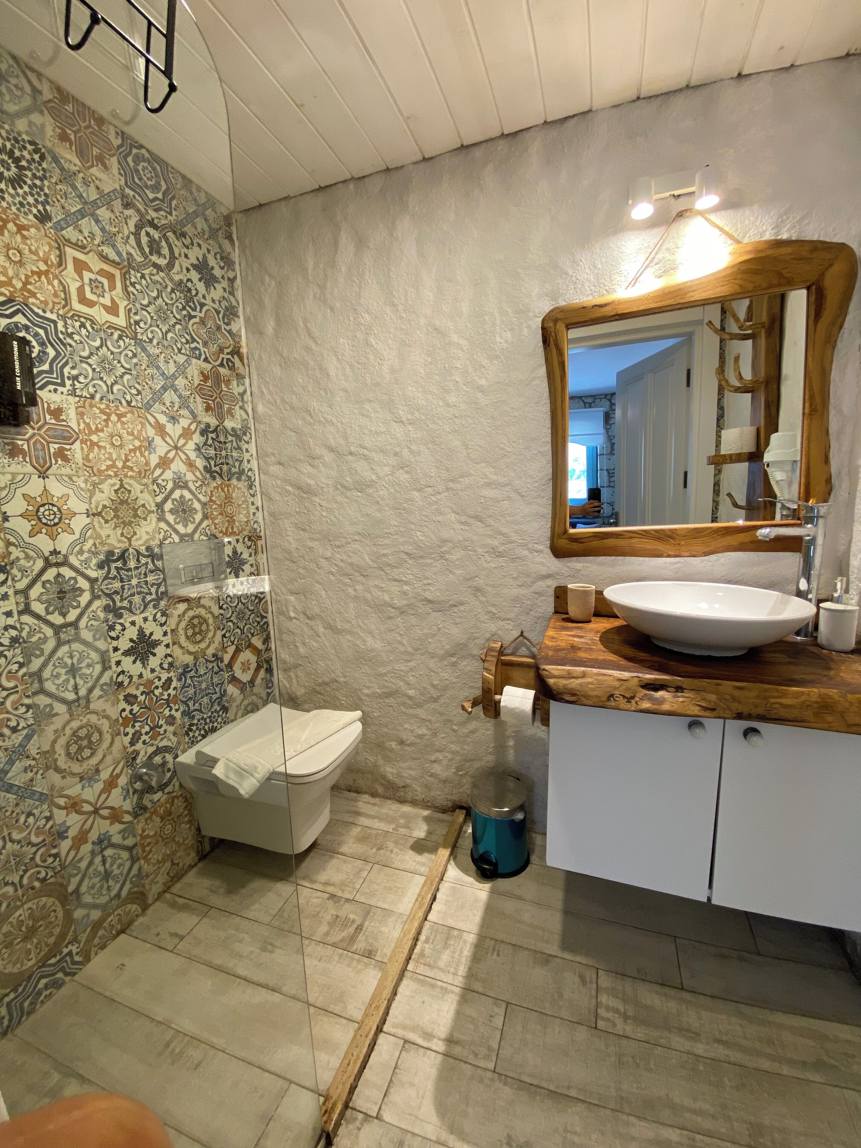 Um banheiro com parede revestida de azulejos estampados, um lavatório de madeira com pia, vaso sanitário e espelho.