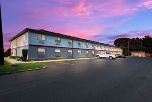 Um motel de dois andares com uma fileira de quartos, estacionamento e céu claro.