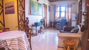 Quarto com cama, cadeira, escrivaninha, ventoinha e janela com cortinas.