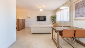 Uma sala de estar moderna com sofá, mesa de jantar e cadeiras. Há uma televisão de tela plana na parede e um vaso com planta.